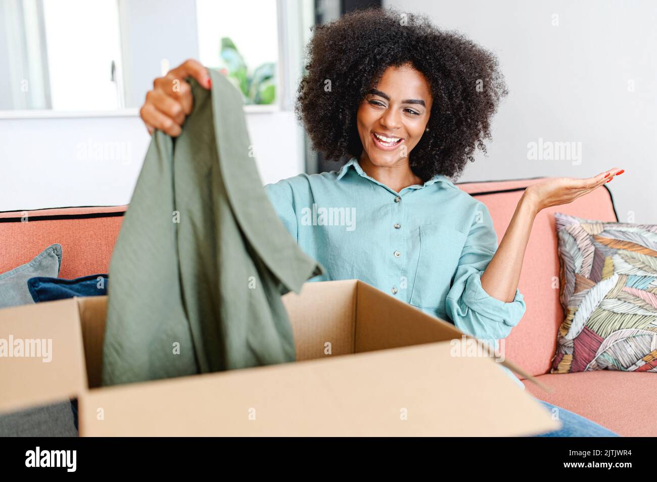 Satisfied 20s young woman sits on the sofa and holding new apparel from ...