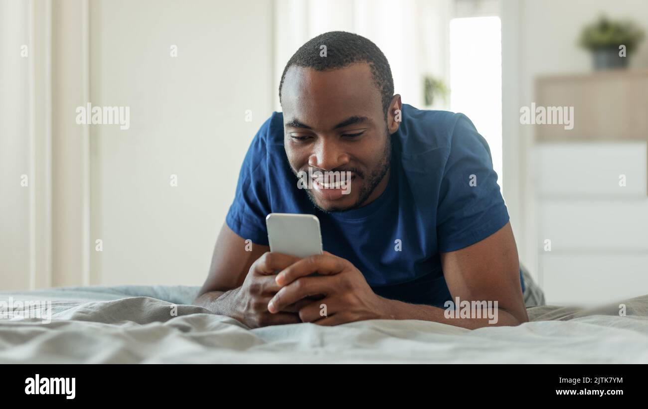 African Guy Texting On Smartphone Lying On Bed In Bedroom Stock Photo ...