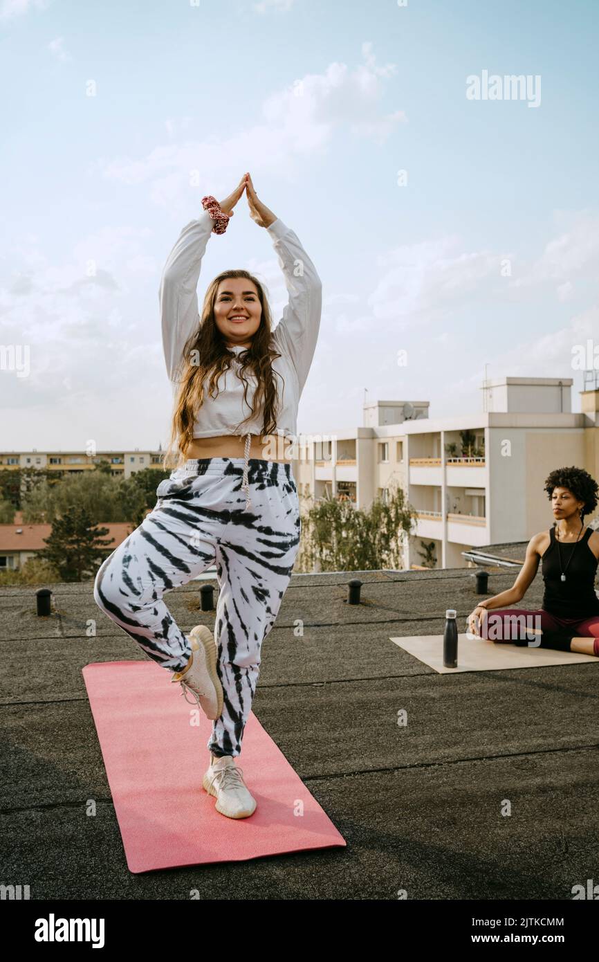 Full length of smiling young woman practicing tree pose by friend ...