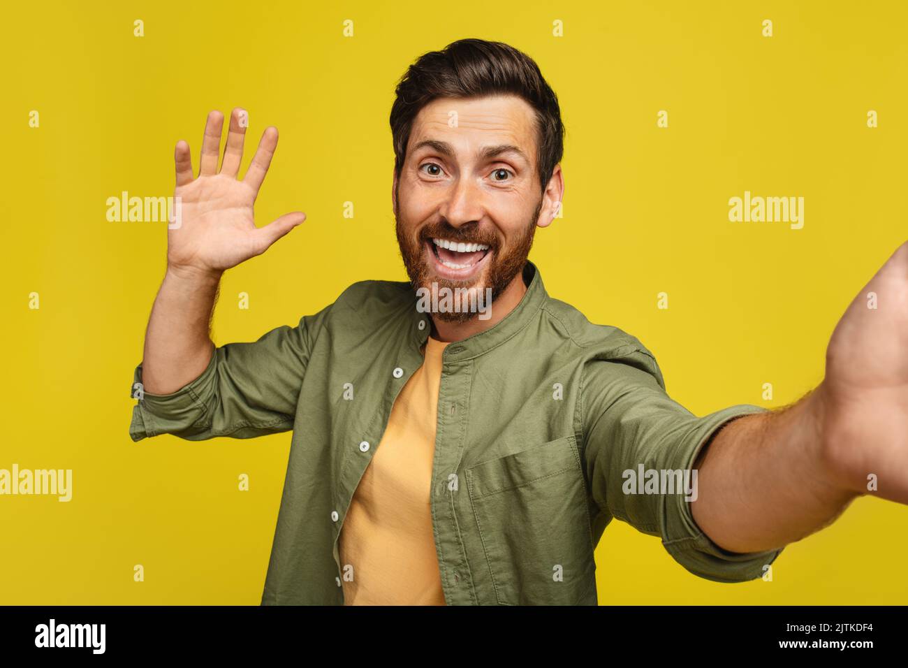 Hello. Excited stylish man taking selfie and gesturing hi at camera over yellow studio ...