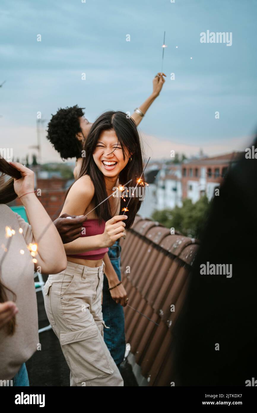 Cheerful young woman holding lit sparkler enjoying with friends on ...