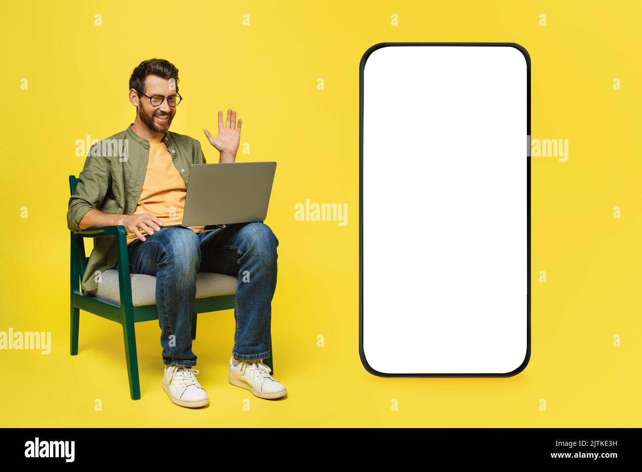 Positive man using laptop, sitting on chair near giant cellphone with ...