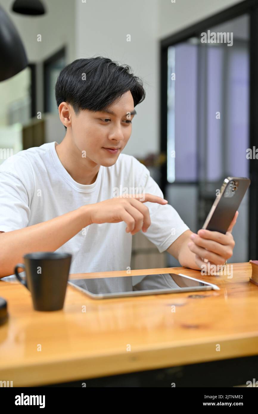 Portrait, Handsome and happy young asian guy sits at his working desk, using his smartphone to ...
