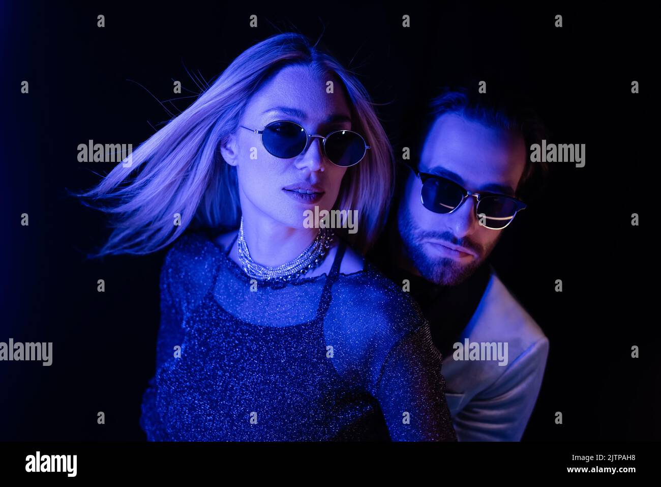 Stylish couple in sunglasses standing isolated on black with blue ...