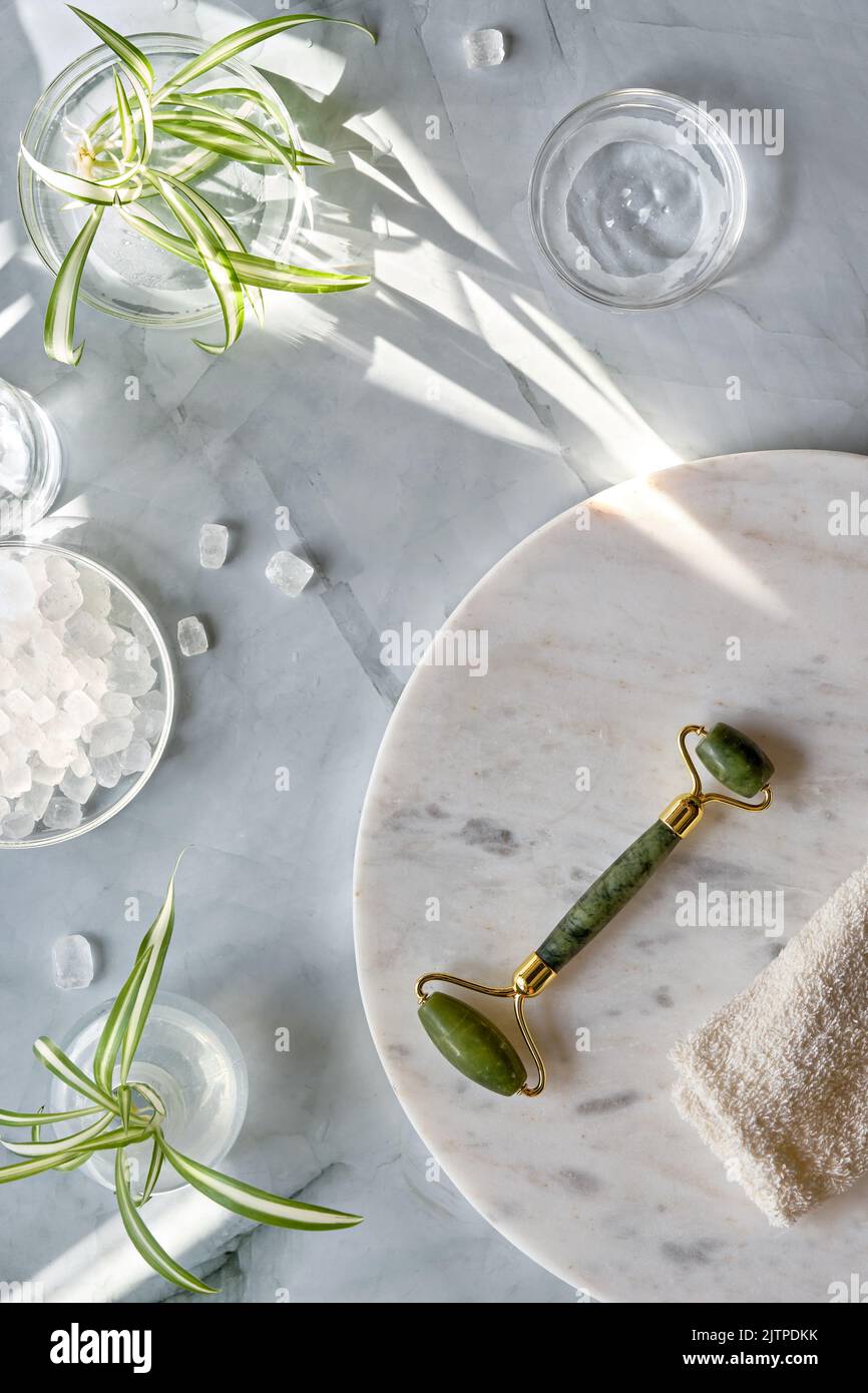 Home made moisturizer and green jade stone face roller on off white marble background. Beauty ...