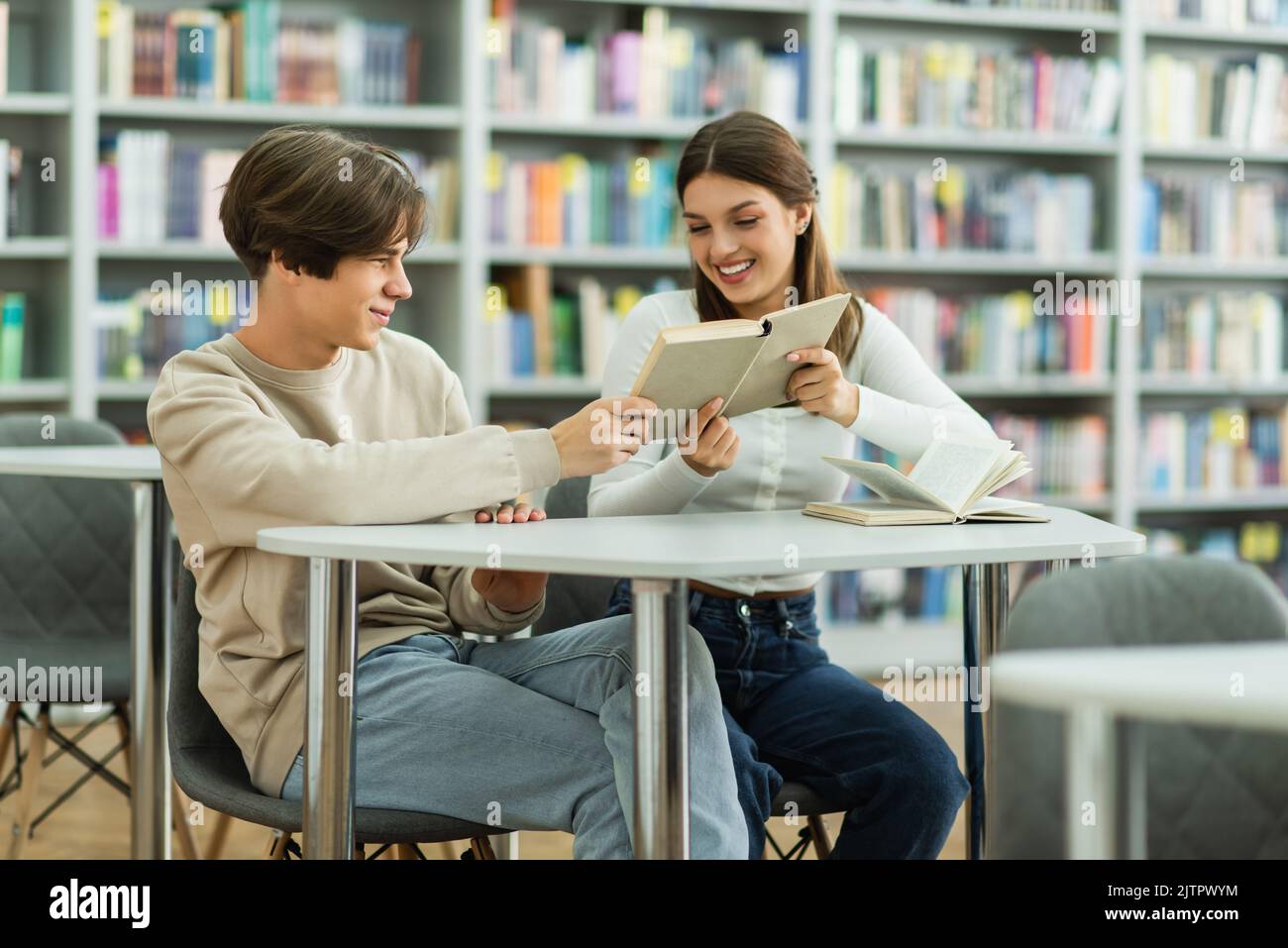 cheerful teen girl showing book to smiling friend in library reading ...