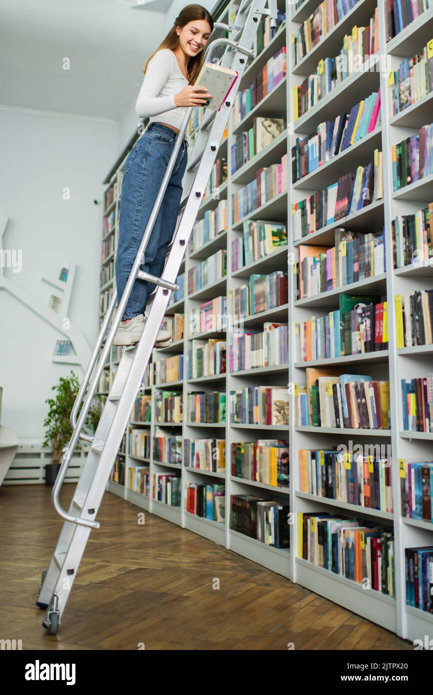 happy teenage girl holding book while standing on ladder in library ...