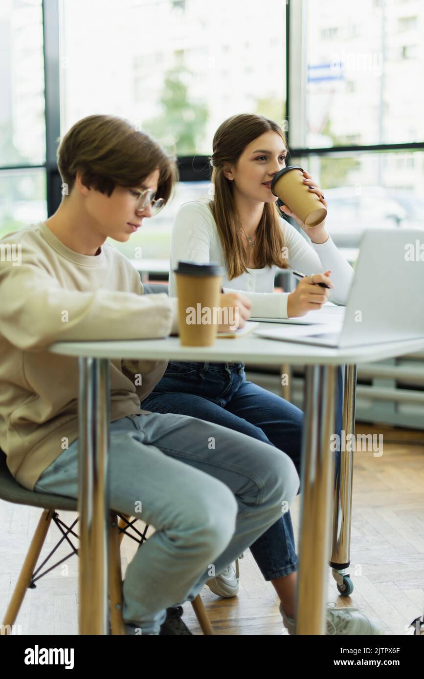 teenage girl drinking coffee near laptop and friend writing in library ...