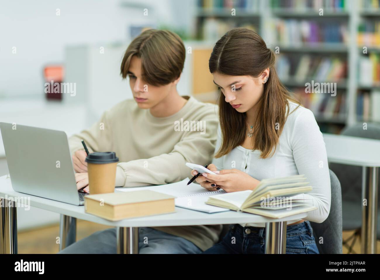 teenage girl using smartphone near student and laptop in library ...