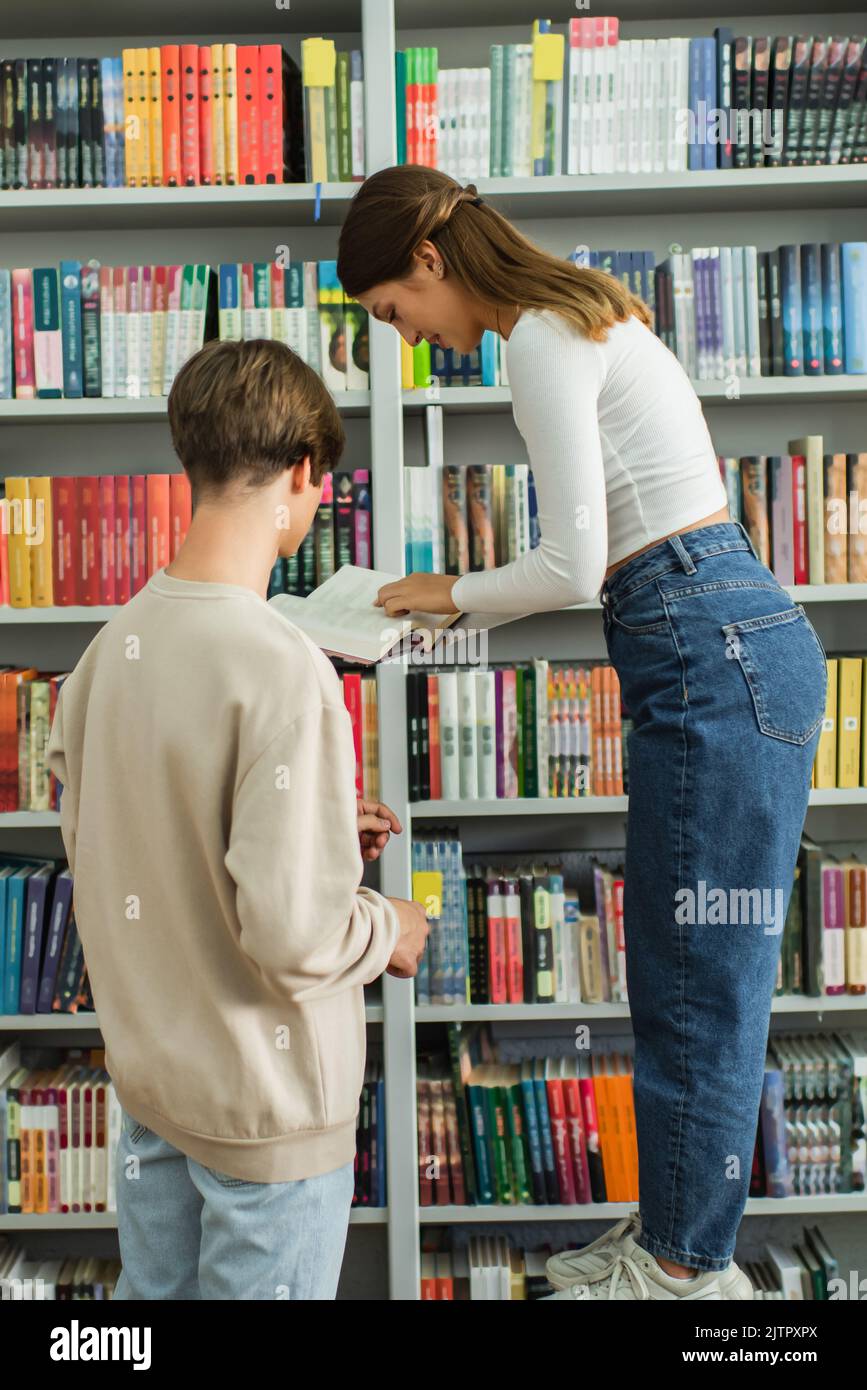 teenage girl showing book to friend near racks in library,stock image ...