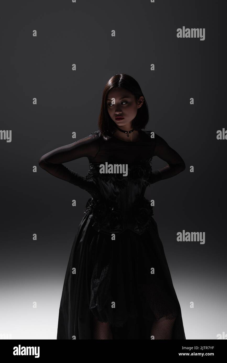 brunette asian model in black gothic dress posing with hands on hips on ...