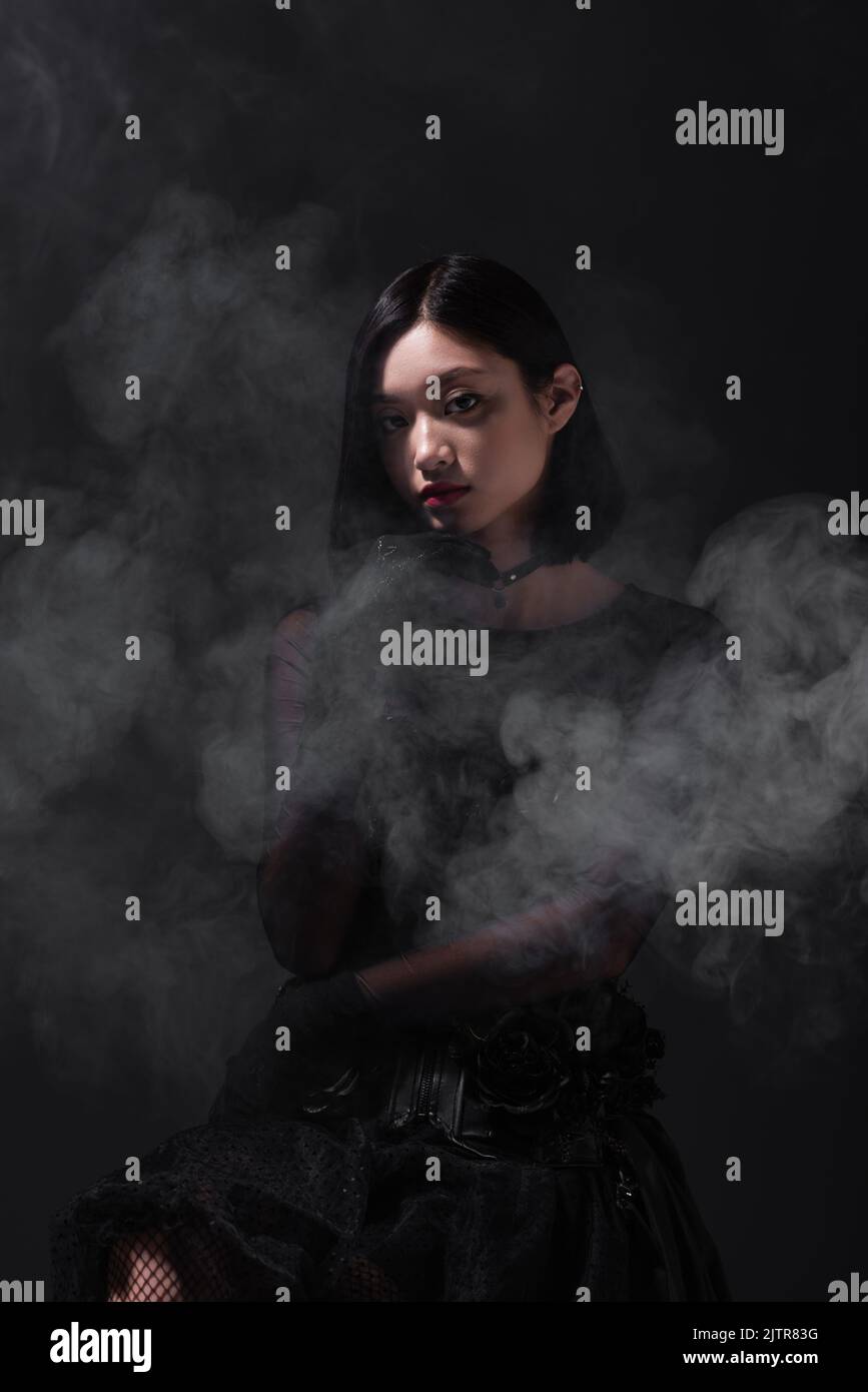 brunette young asian woman in gothic outfit and gloves on grey with ...
