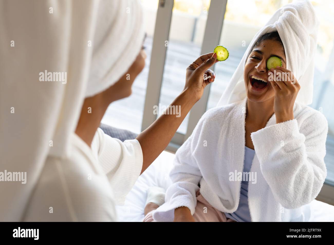 Cheerful biracial daughter and mother putting cucumber slices on eyes ...