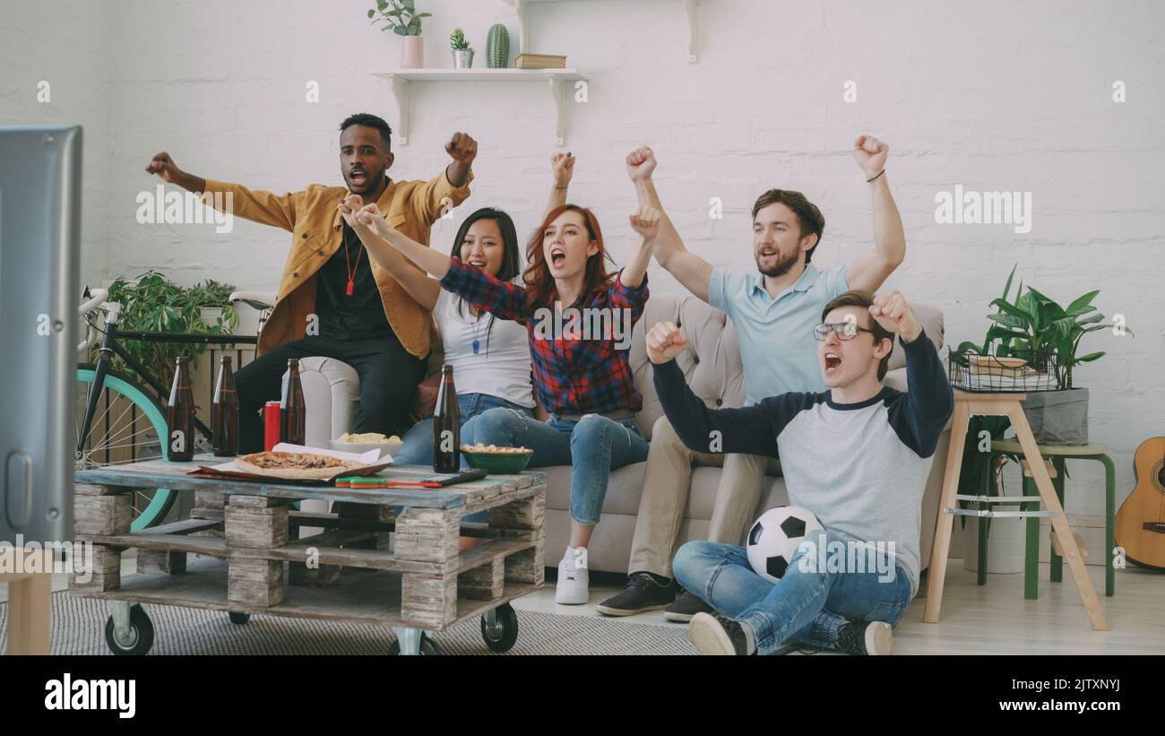 Group of happy friends watching sports game on TV at home indoors. They ...