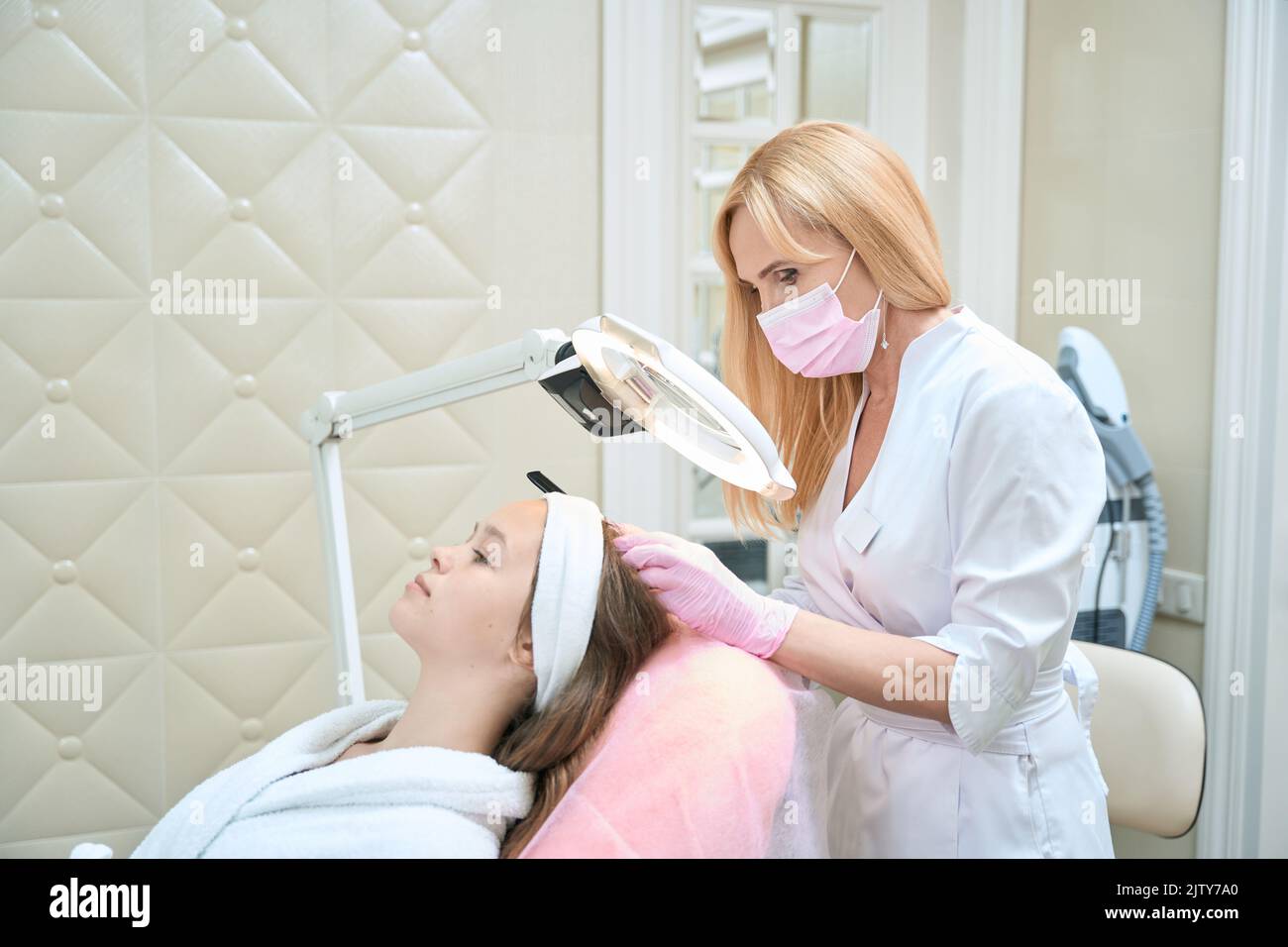 Cosmetologist checks girl head, looking through magnifying glass lamp ...