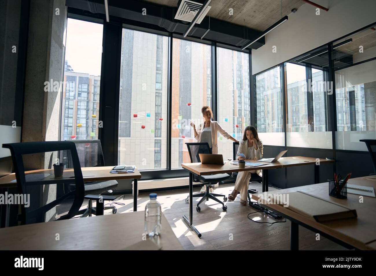 Pretty female colleagues are chatting while being in spacious office ...