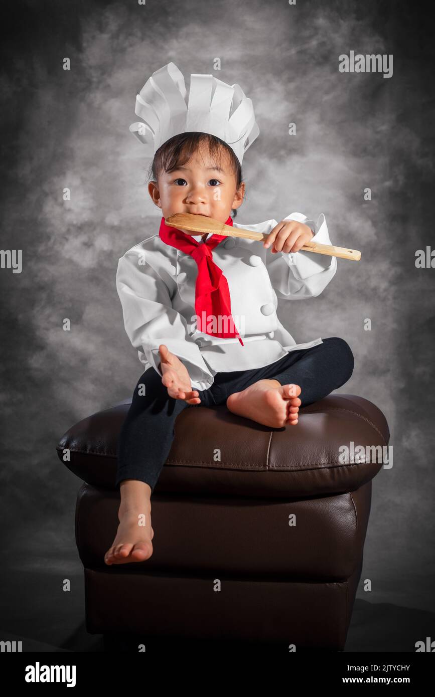 A toddler girl in a chef cook costume portrait is biting a wooden ...