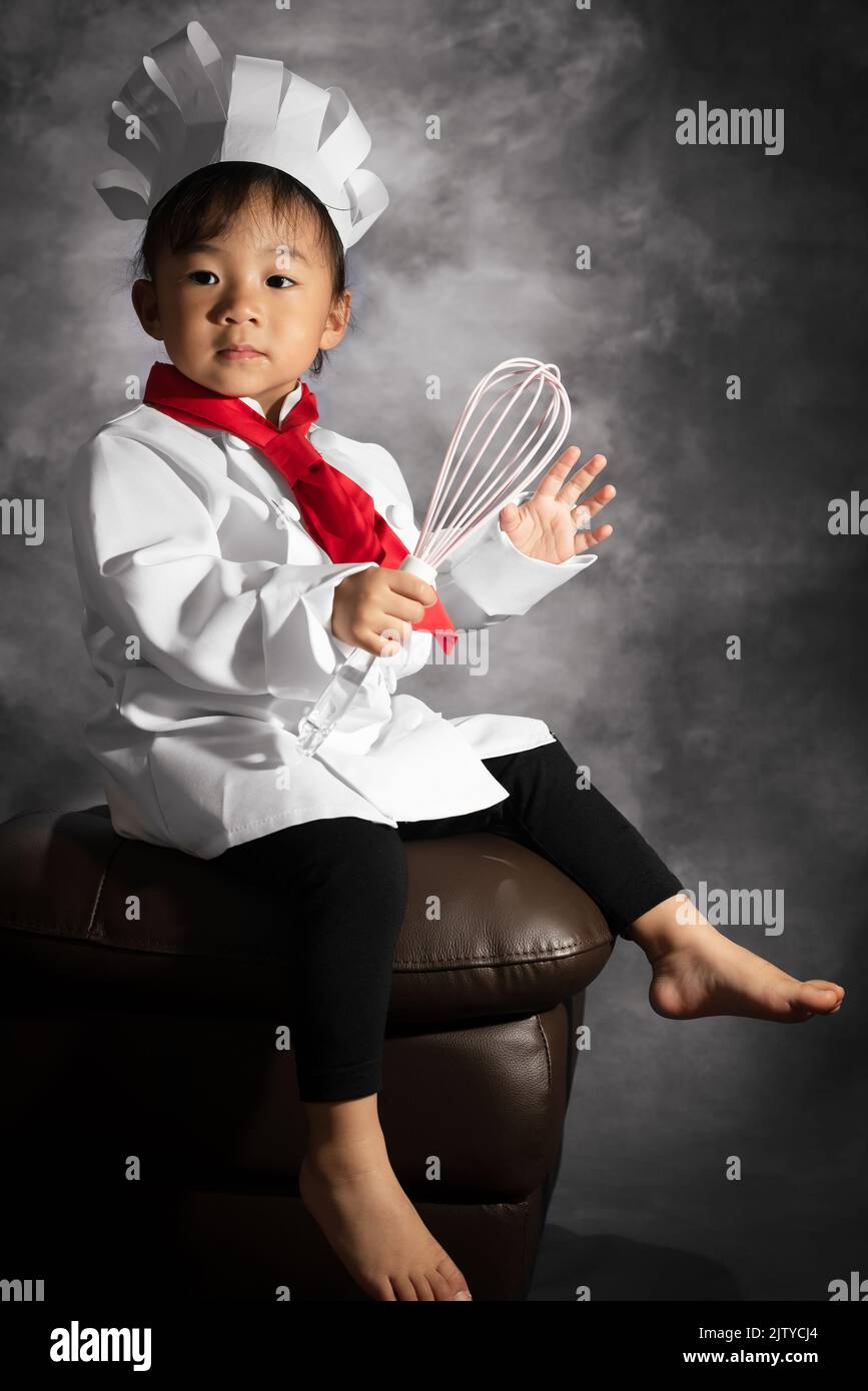 A toddler girl portrait is holding a whisk while sitting on a chair ...