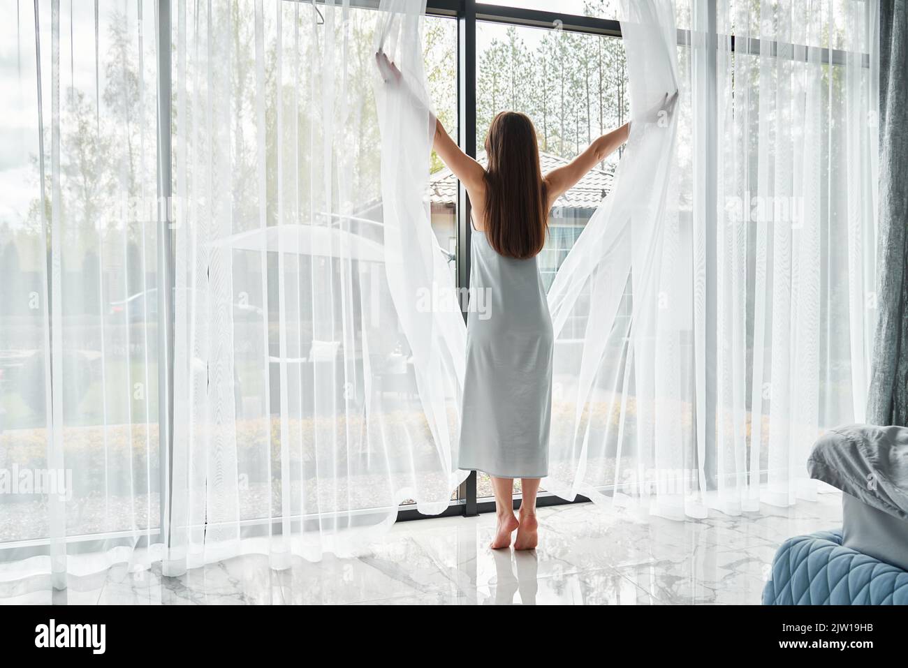 Young woman stand and open white curtains at the window, the morning ...