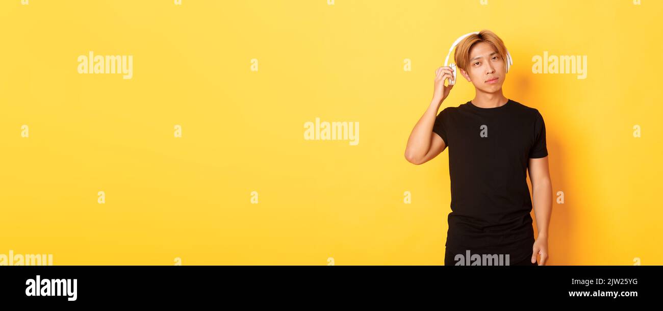 Young handsome asian guy with blond hair, take-off headphones to listen ...