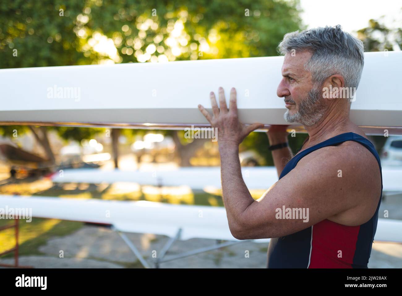 Senior caucasian male rower carrying boat on his shoulders to the lake ...