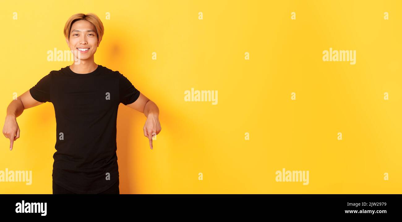 Portrait Of Smiling Attractive Korean Guy With Blond Hair Wearing Black Outfit Pointing