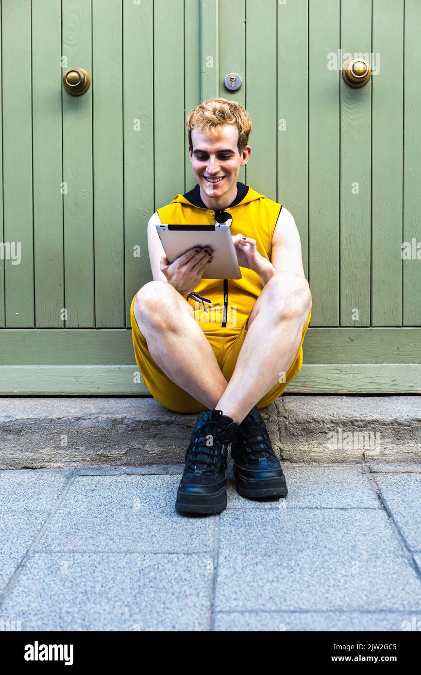 Young smiling transgender man in yellow jumpsuit browsing tablet while ...