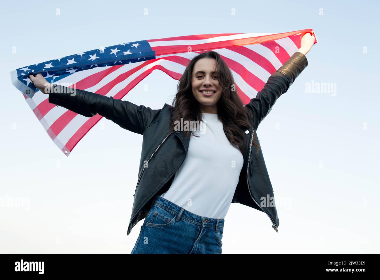 Smiling young caucasian woman waving american flag against the blue sky ...