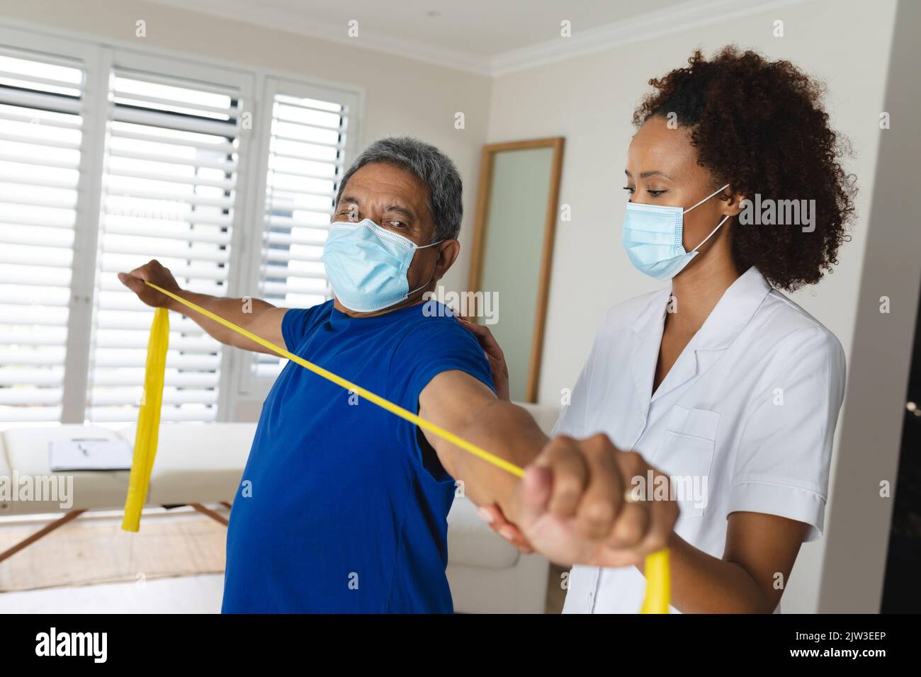 Mixed race female physiotherapist wearing mask helping senior exercise ...