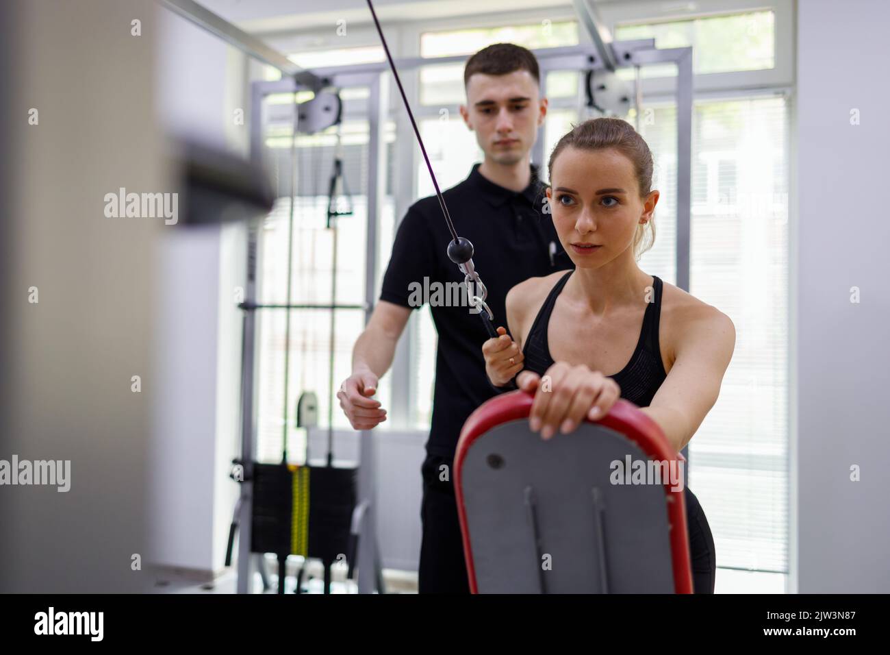 Physical instructor assisting young woman working out on pull cable ...
