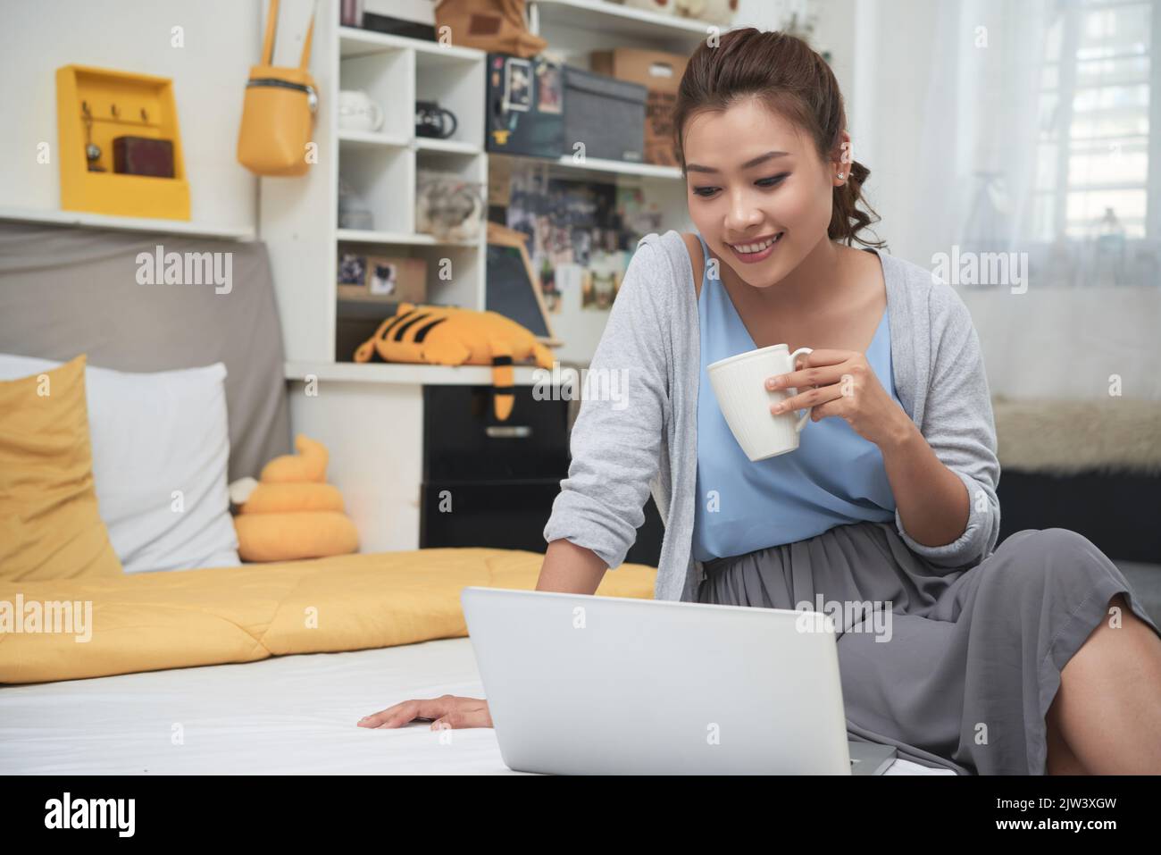 Pretty young Vietnamese woman sitting on bed in her room and working on ...