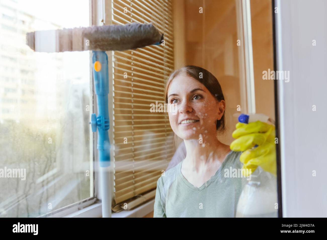 Cheerful young woman cleaning window with squeegee and spray detergent ...