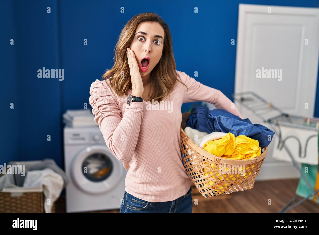 Young woman holding laundry basket afraid and shocked, surprise and amazed expression with hands ...