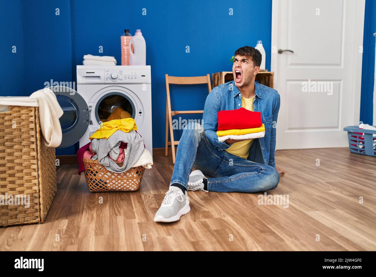 Young hispanic man holding clean laundry angry and mad screaming frustrated and furious ...