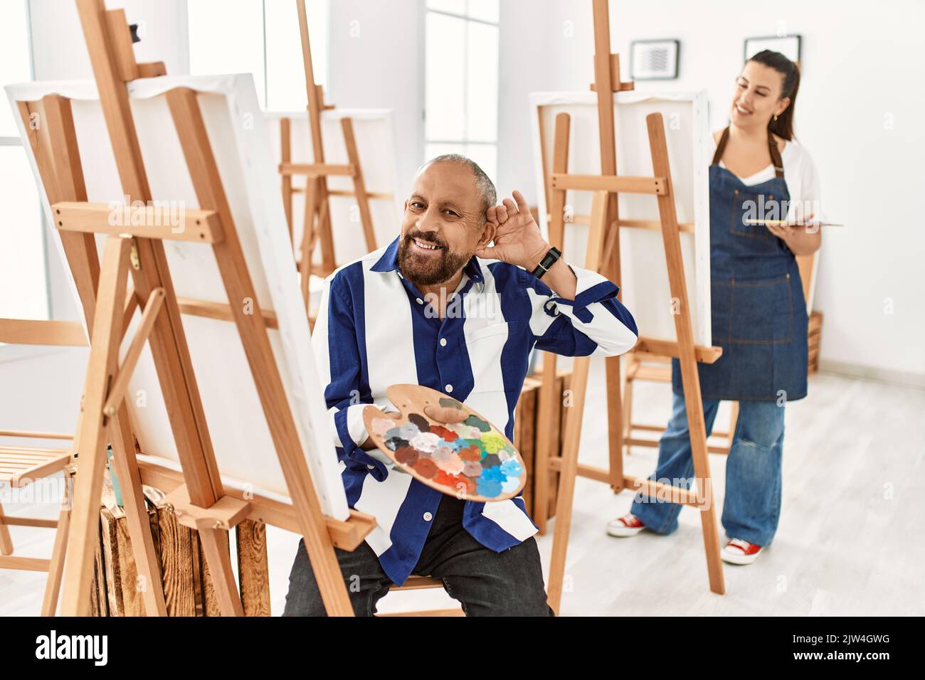 Senior artist man at art studio smiling with hand over ear listening an ...