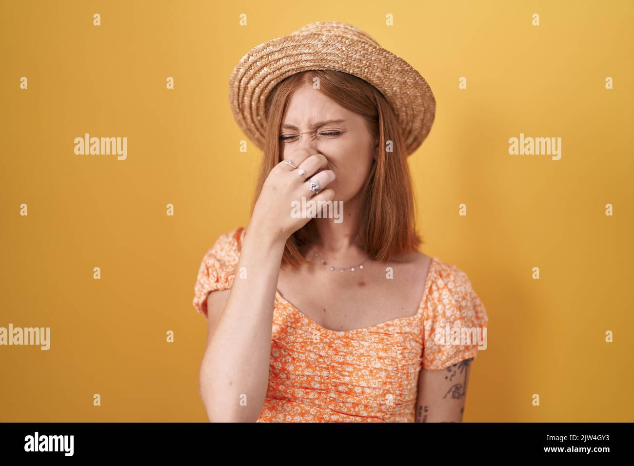 Young redhead woman standing over yellow background wearing summer hat ...