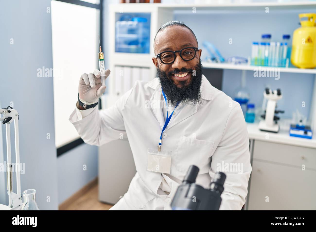 African american man working at scientist laboratory holding syringe looking positive and happy ...