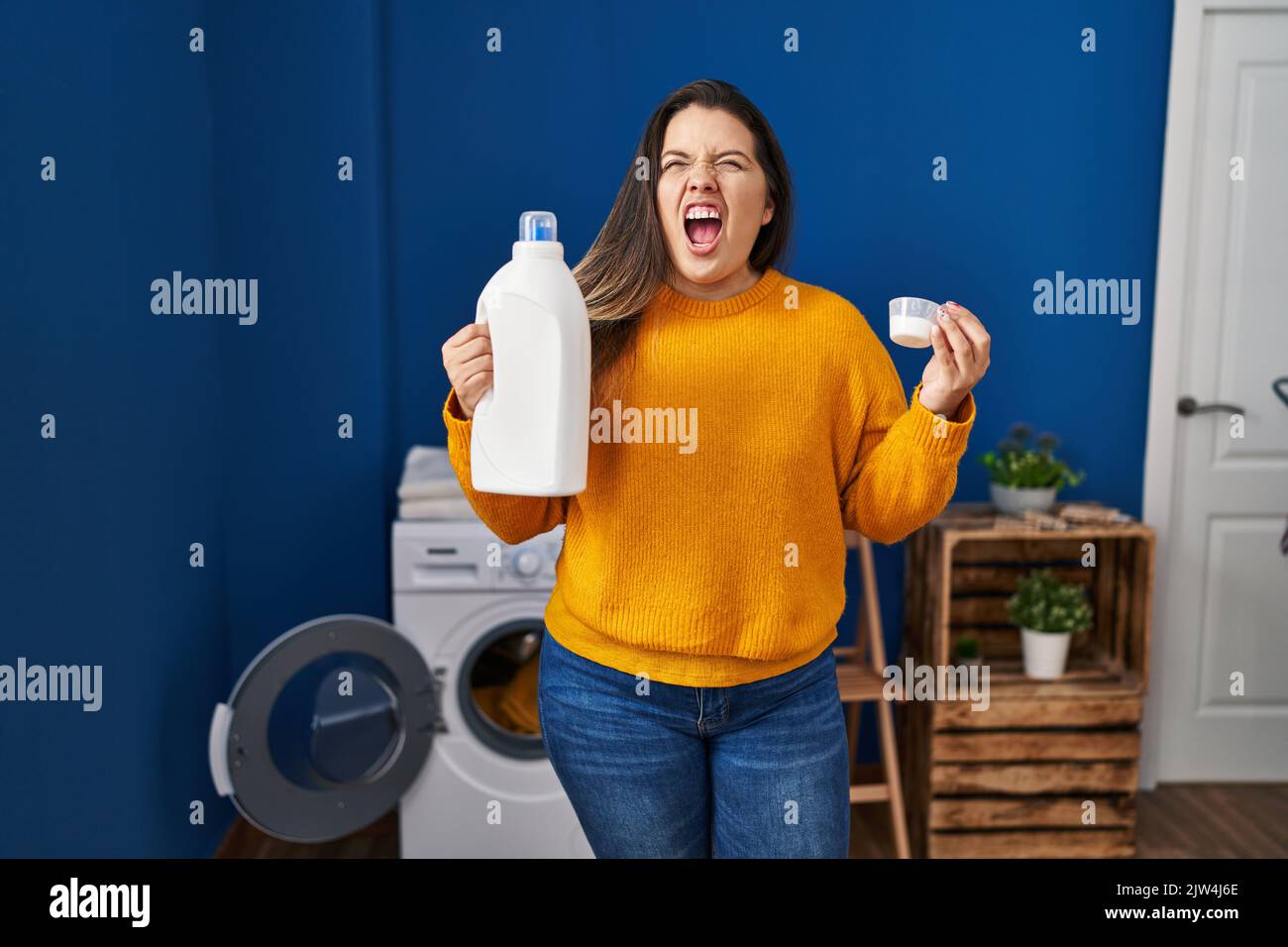 Young hispanic woman holding detergent bottle angry and mad screaming ...