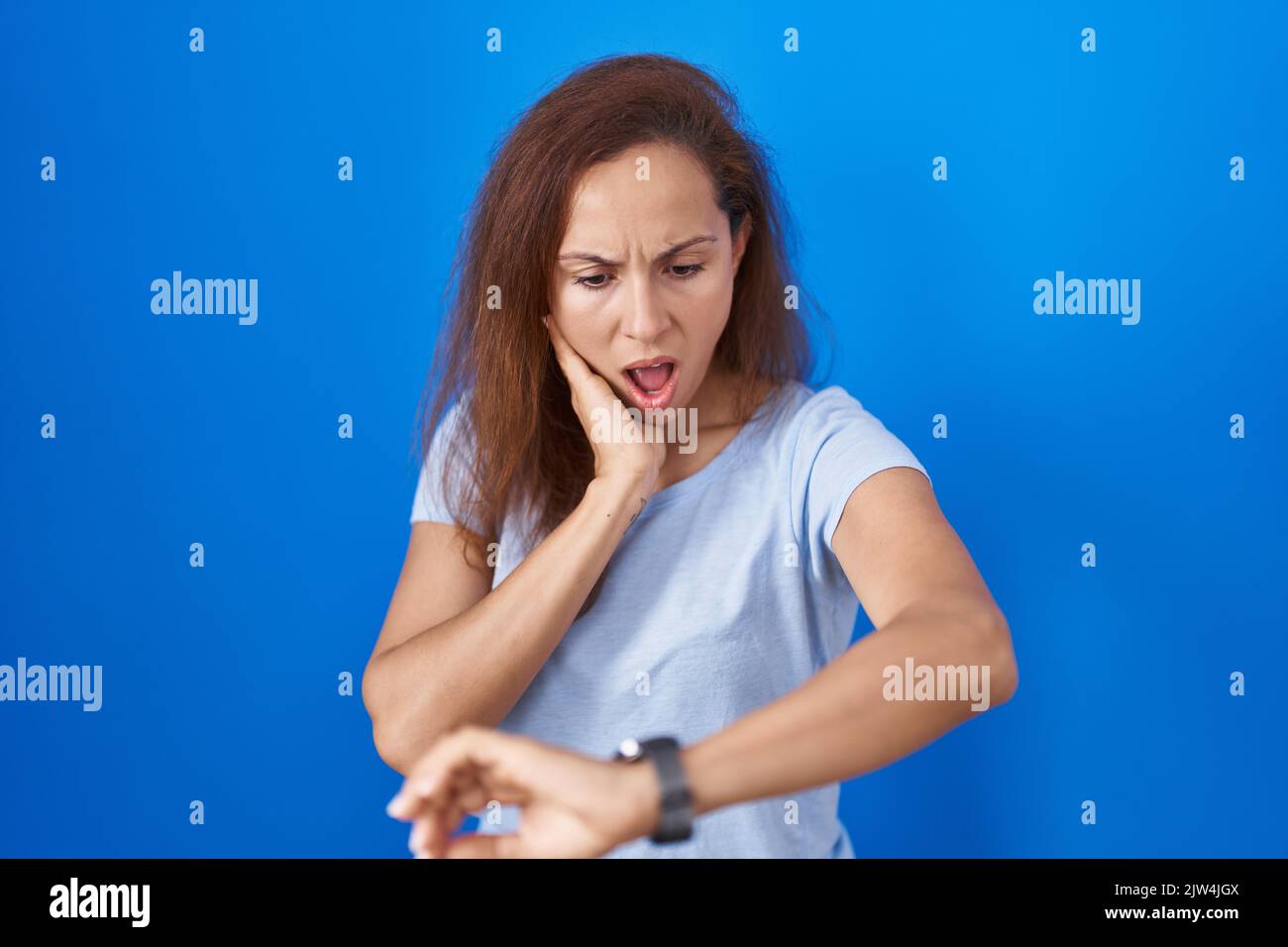 Brunette woman standing over blue background looking at the watch time ...