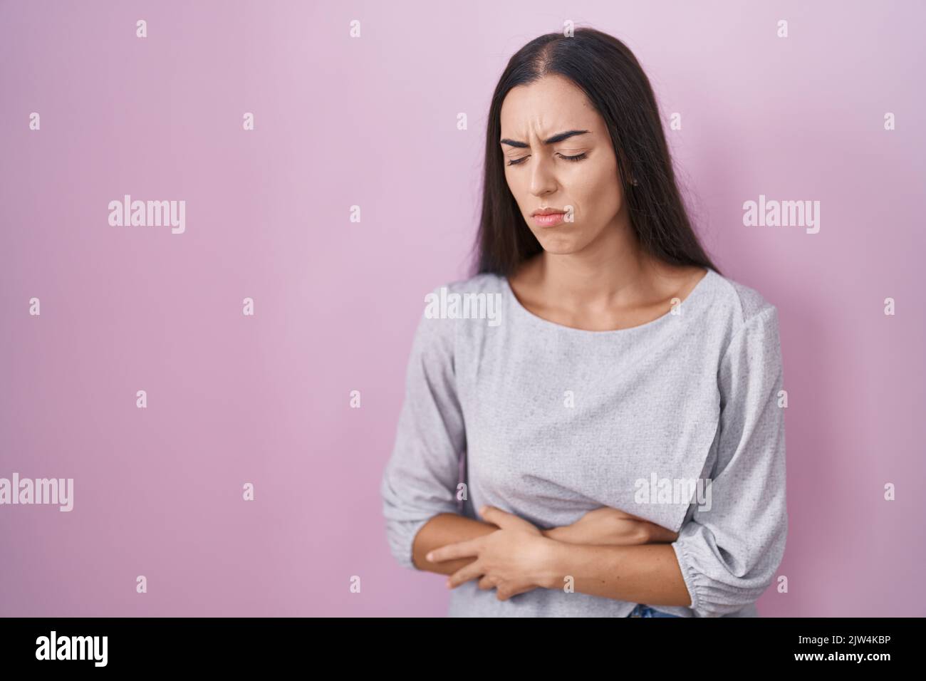 Young brunette woman standing over pink background with hand on stomach ...