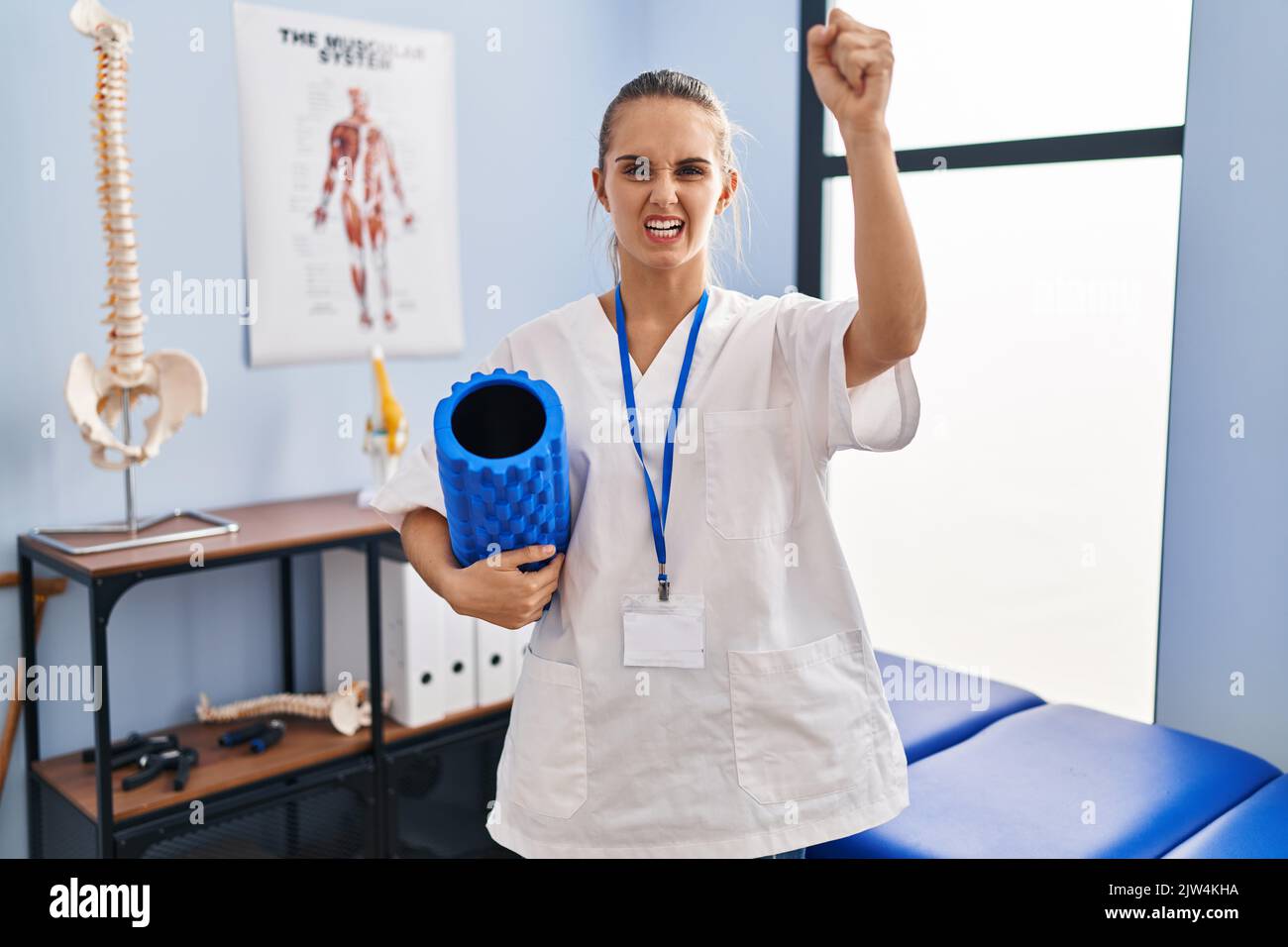 Young physiotherapist woman holding foam roll at the clinic annoyed and ...