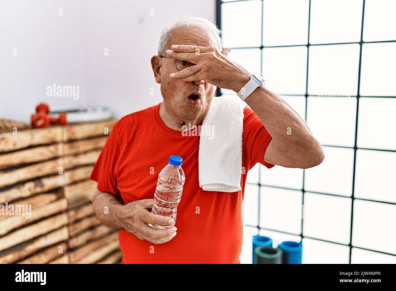 Senior man wearing sportswear and towel at the gym peeking in shock ...