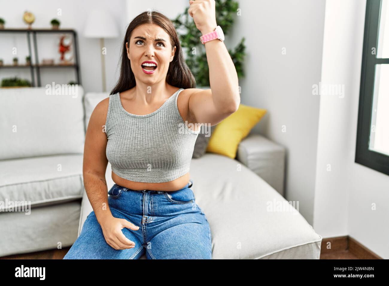 Young brunette woman sitting on the sofa at home angry and mad raising ...