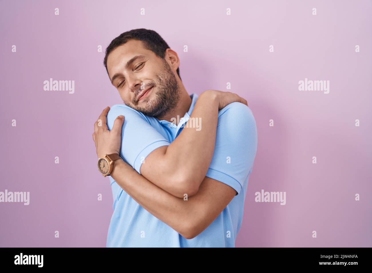 Hispanic man standing over pink background hugging oneself happy and ...