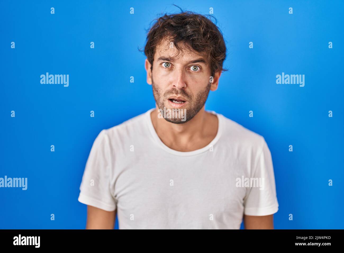 Hispanic young man standing over blue background in shock face, looking ...