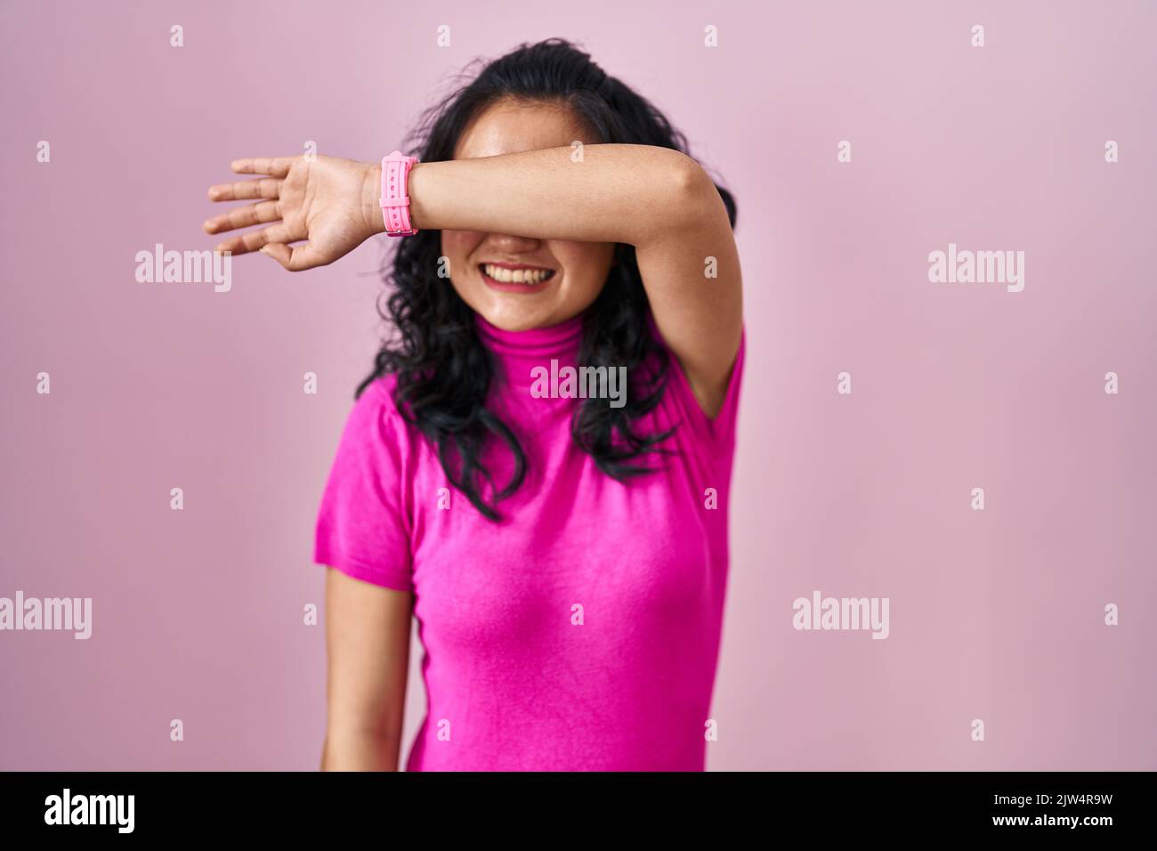 Young asian woman standing over pink background covering eyes with arm ...