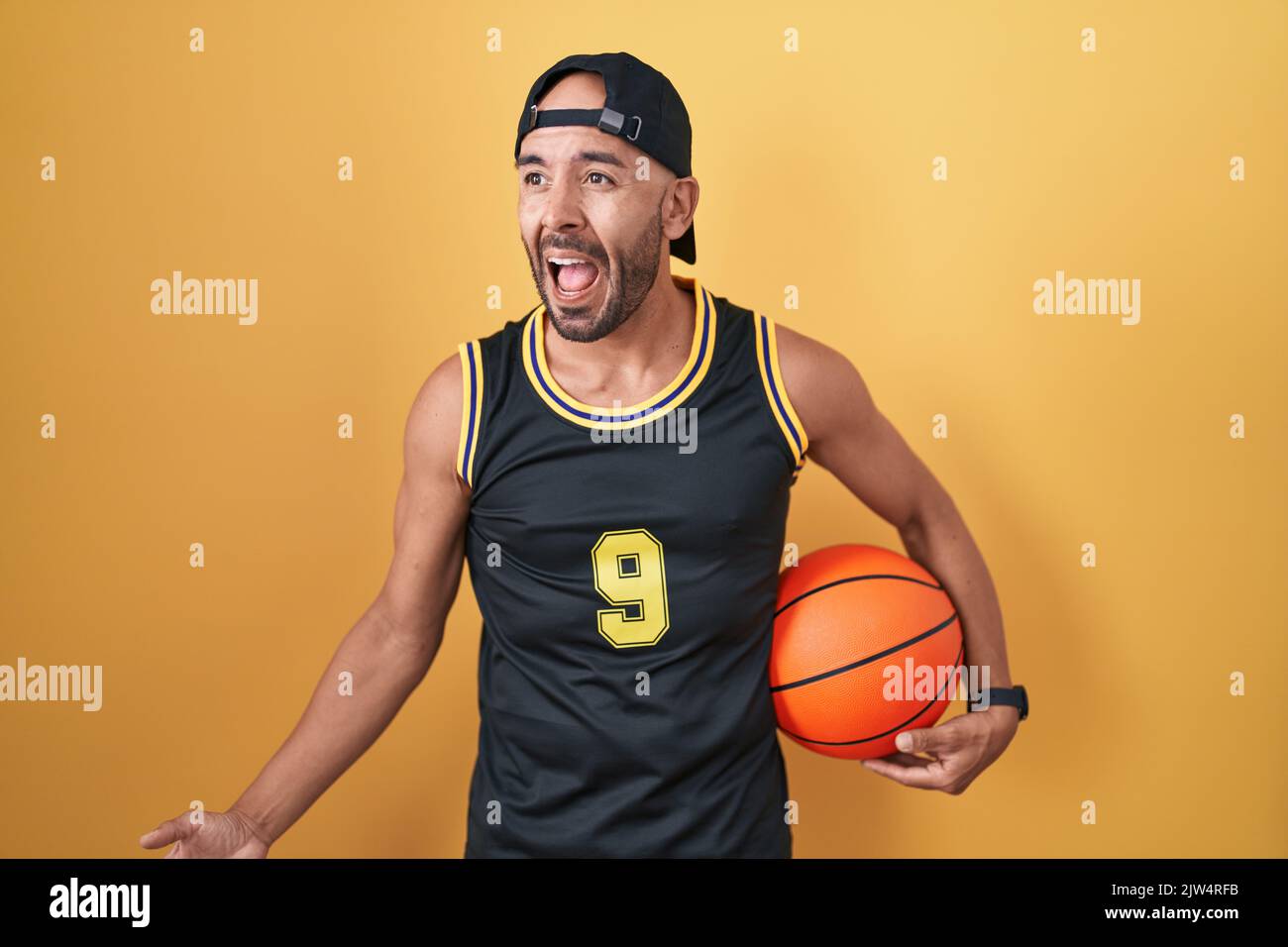 Middle age bald man holding basketball ball over yellow background ...