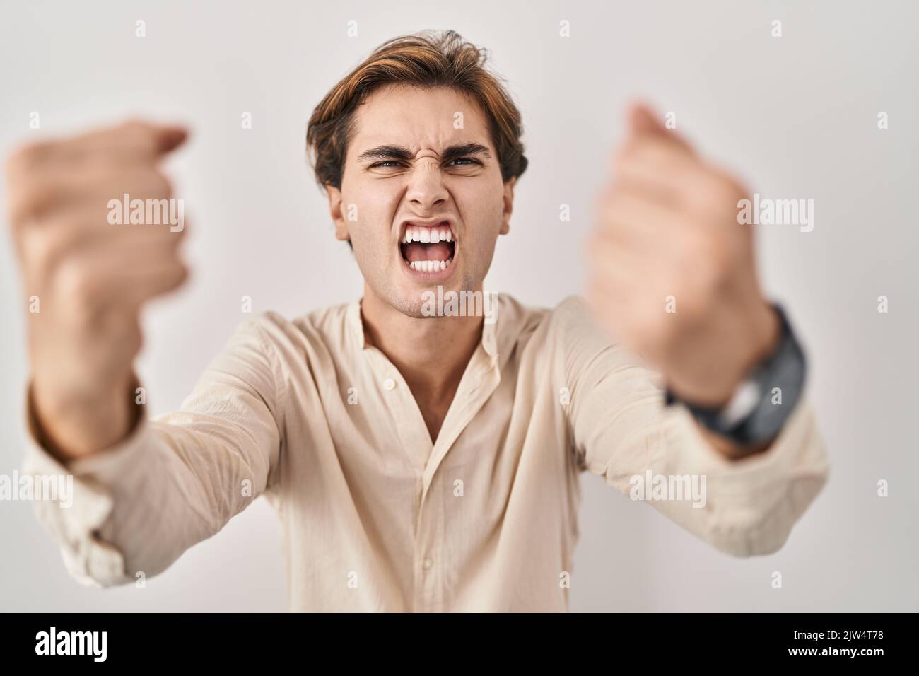 Young man standing over isolated background angry and mad raising fists ...