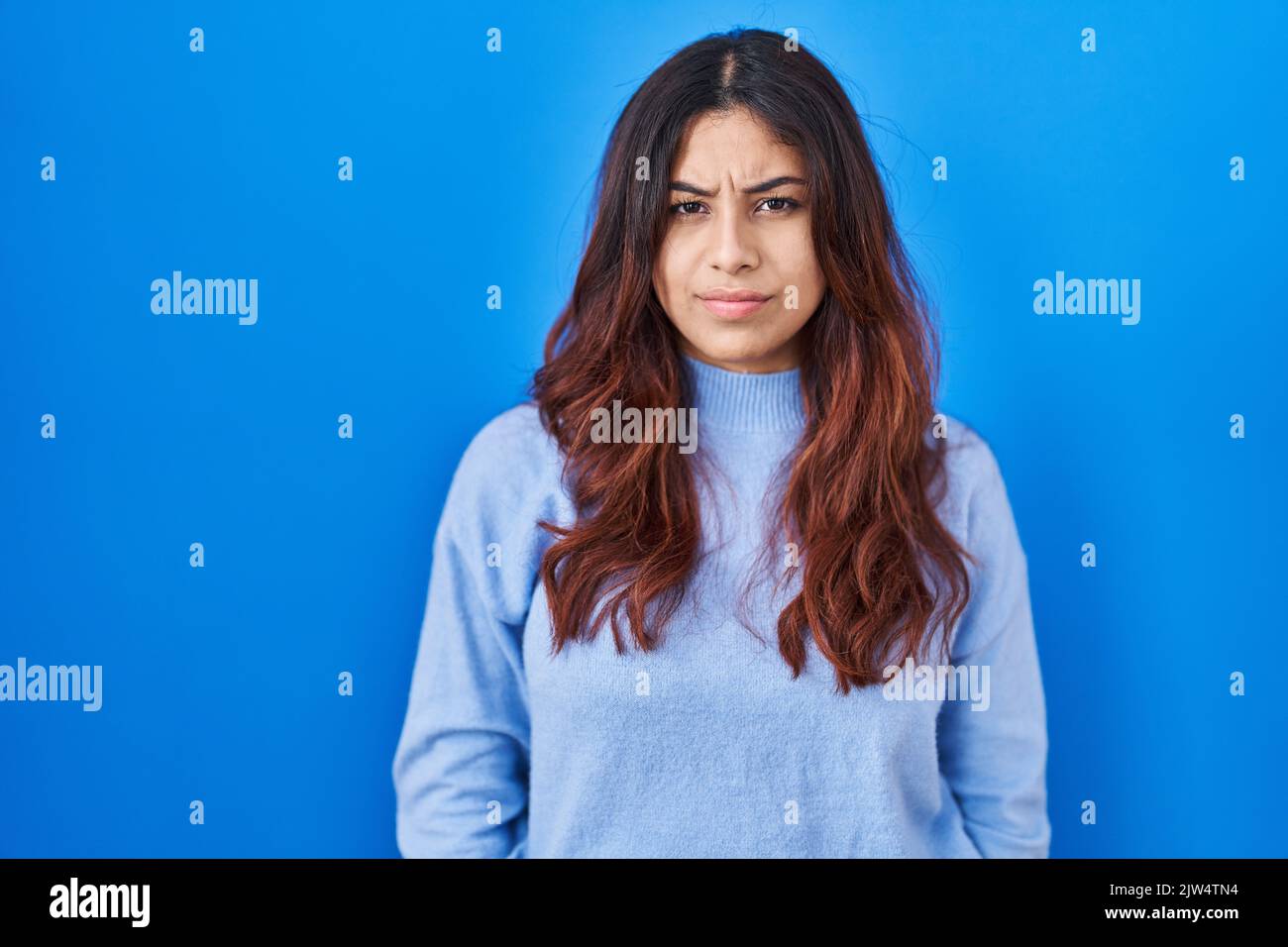 Hispanic young woman standing over blue background skeptic and nervous ...