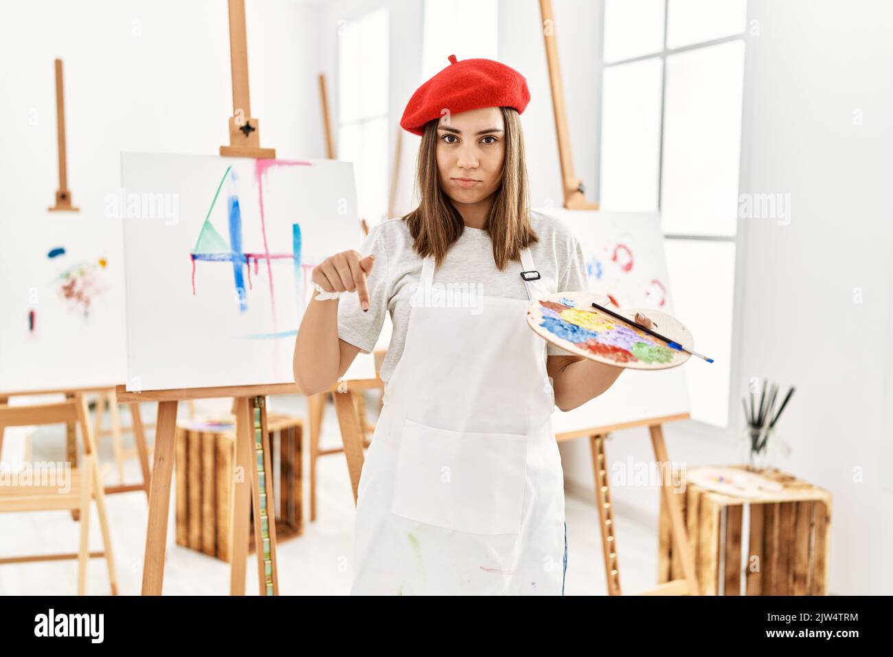 Young artist woman painting on a canvas at art studio pointing down ...